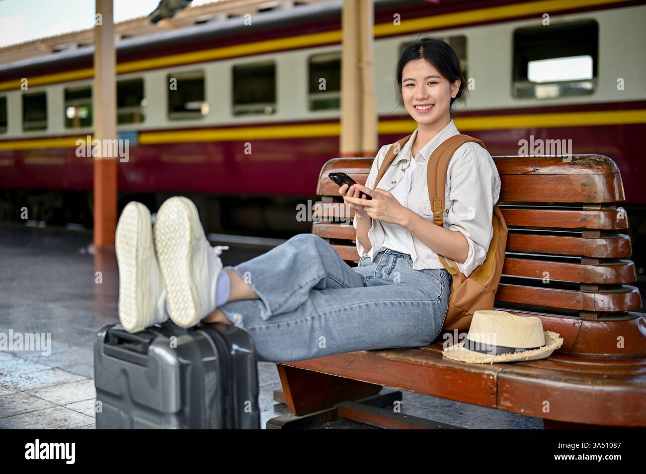 Smiling Asian female tourist holding smartphone sitting on bench with ...