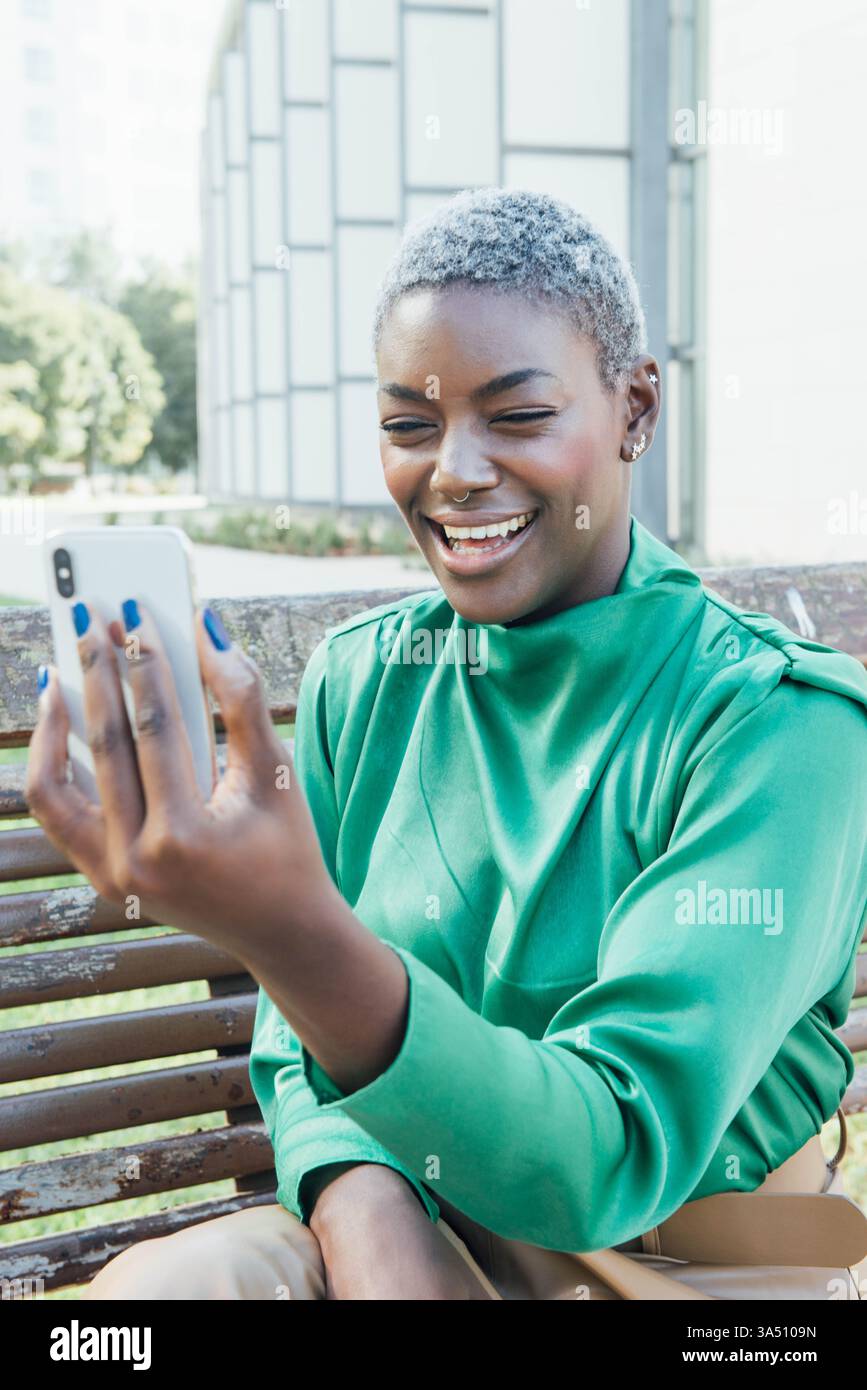 Smiling Black woman having video call sitting sideways on bench in park ...