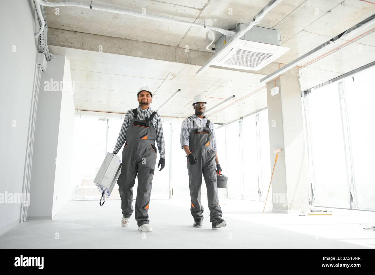 Diverse group of male builders wearing uniform and hard hats carrying tools walking on ...