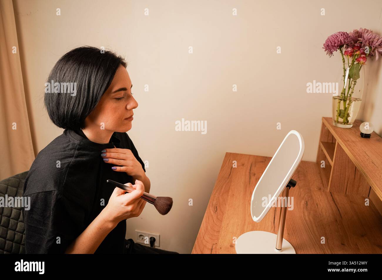 Focusing on her makeup, the woman uses a soft brush to enhance her complexion. Her delicate ...