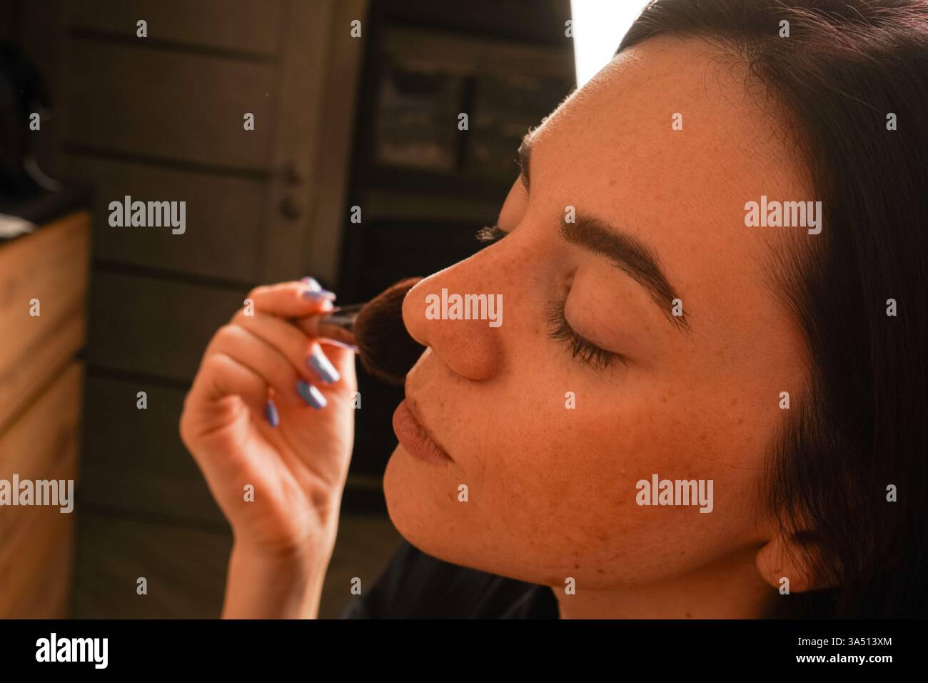 A young woman gently applies powder to her face using a makeup brush ...