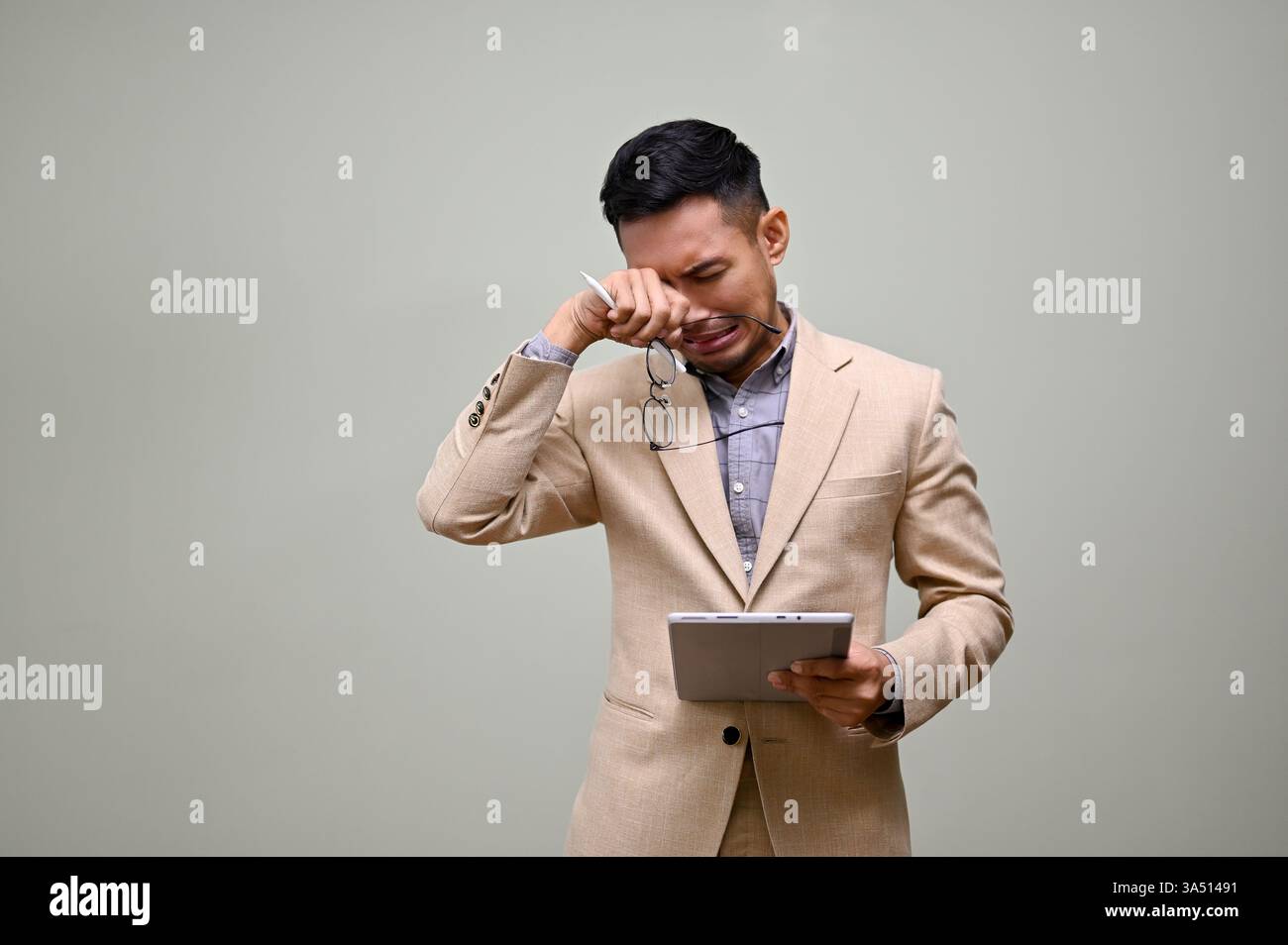 Crying Asian businessman with closed fist on head holding tablet ...