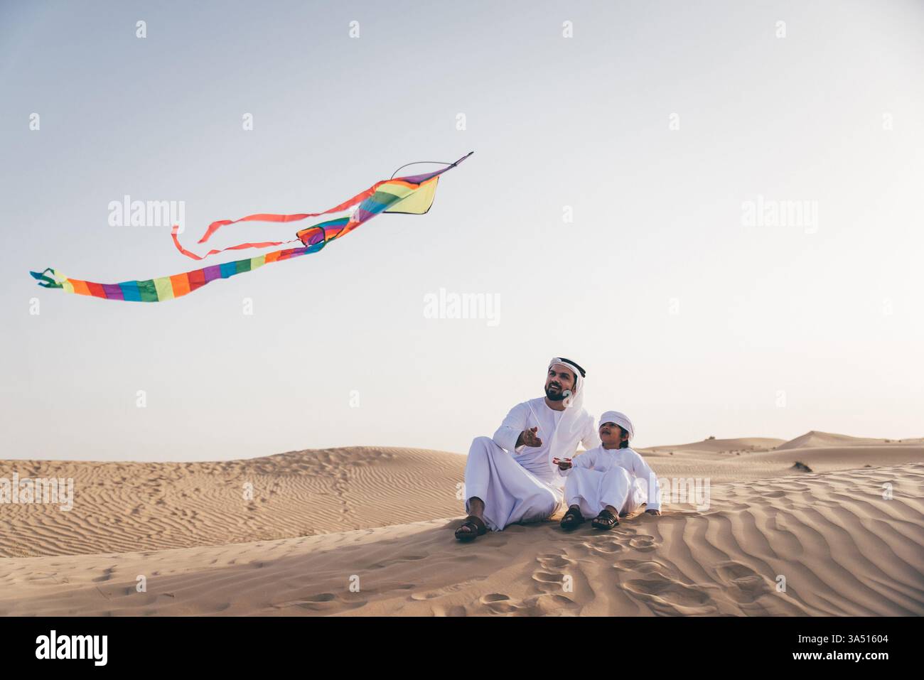 Cheerful Middle Eastern man and son in traditional clothing sitting on ...