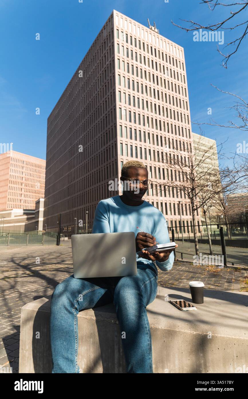 Black man taking notes while working with laptop outside university ...