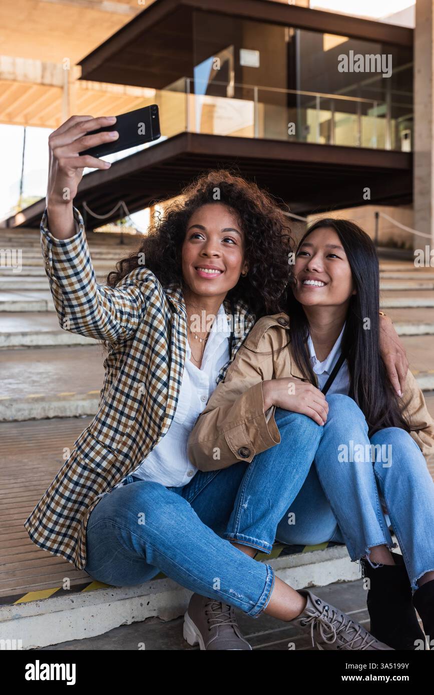 Hispanic and Asian women taking selfie while sitting close together on ...
