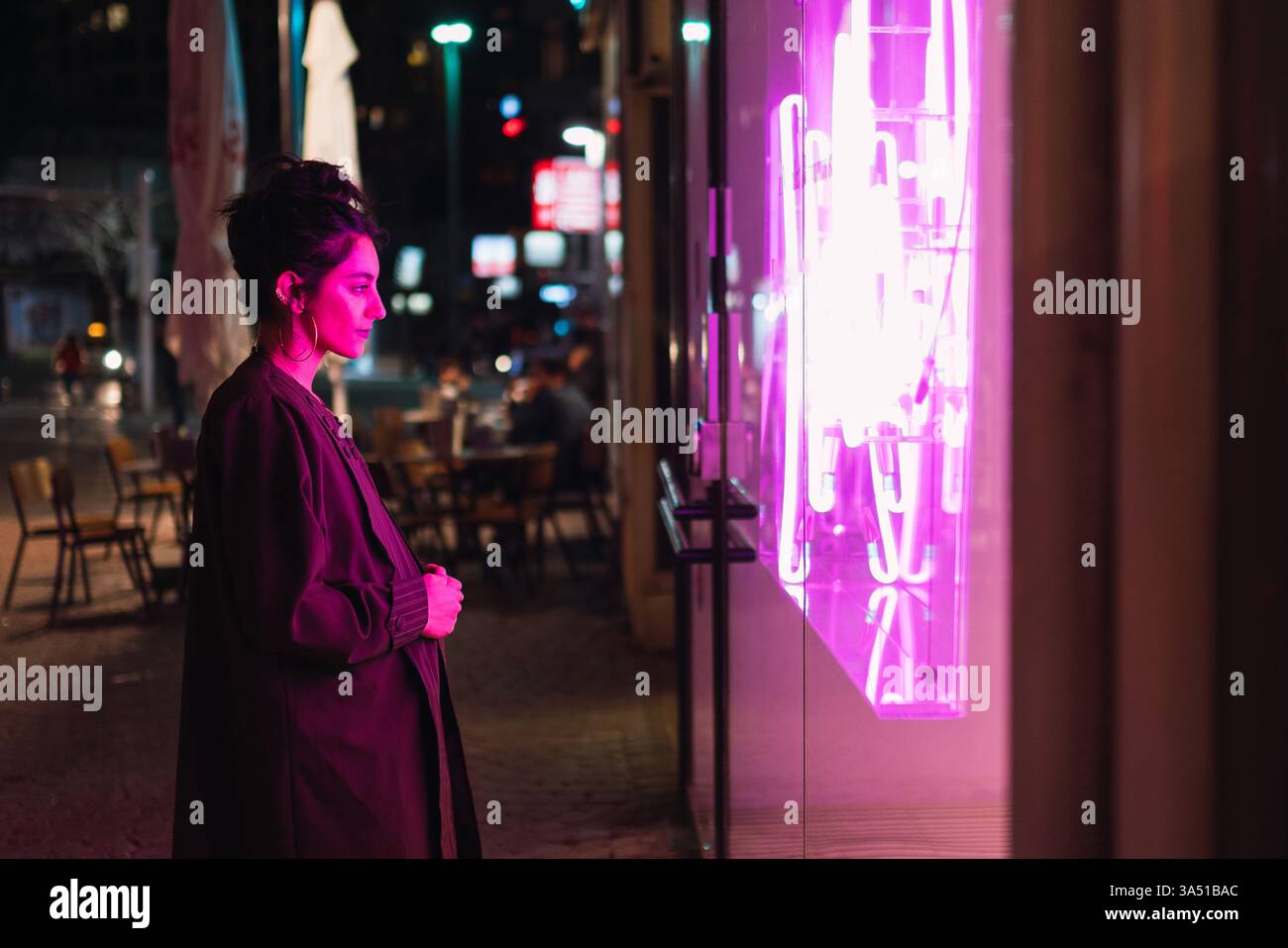 Middle Eastern woman gazes at a neon-lit wall on a city street at night ...