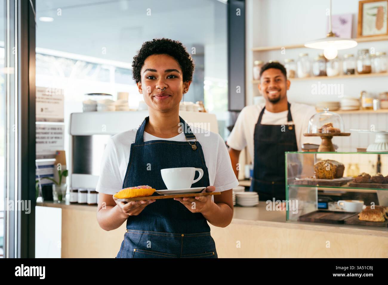 Smiling mixed-race female barista wearing apron holding tray with ...