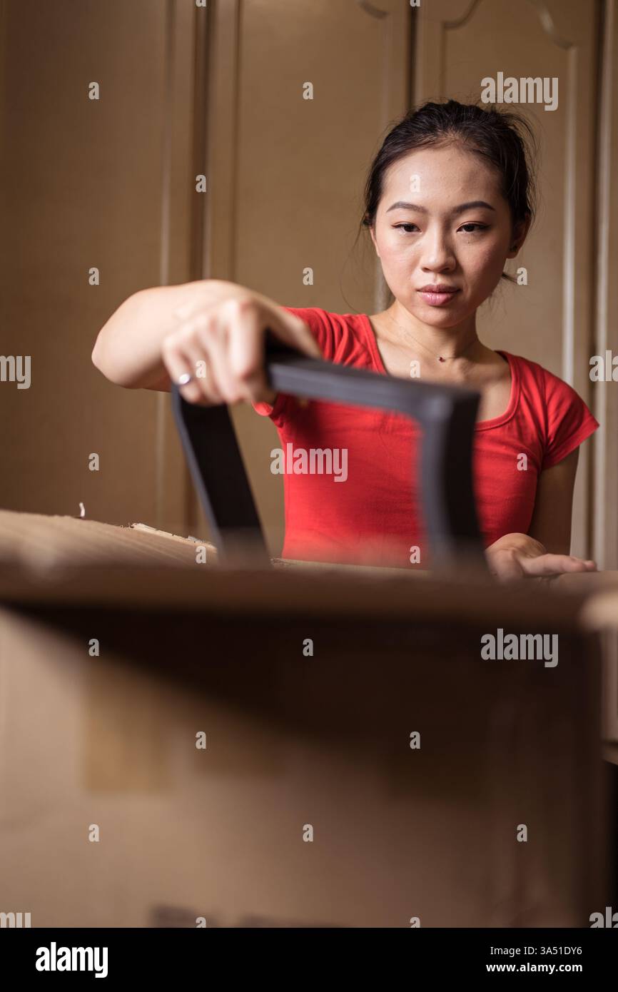 Asian woman unpacking carton box with disassembled chair parts at home ...