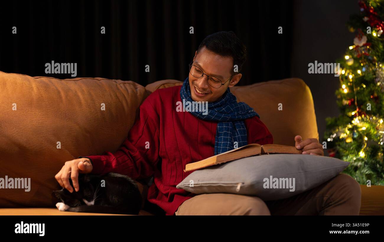 A happy Asian man in a cosy sweater and scarf is petting his cute cat ...