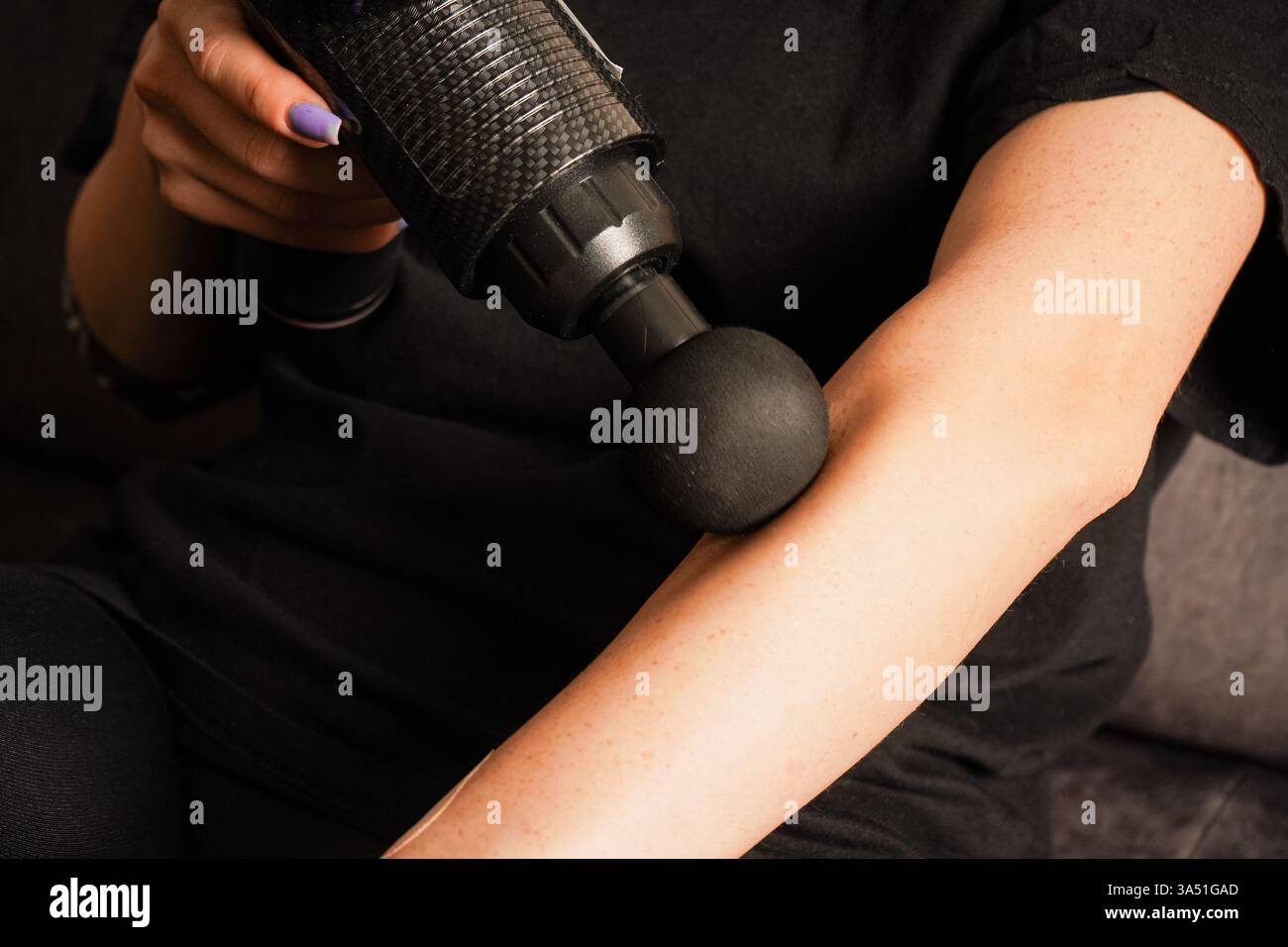 Relaxing indoors, a woman applies a massage gun to her leg for recovery ...