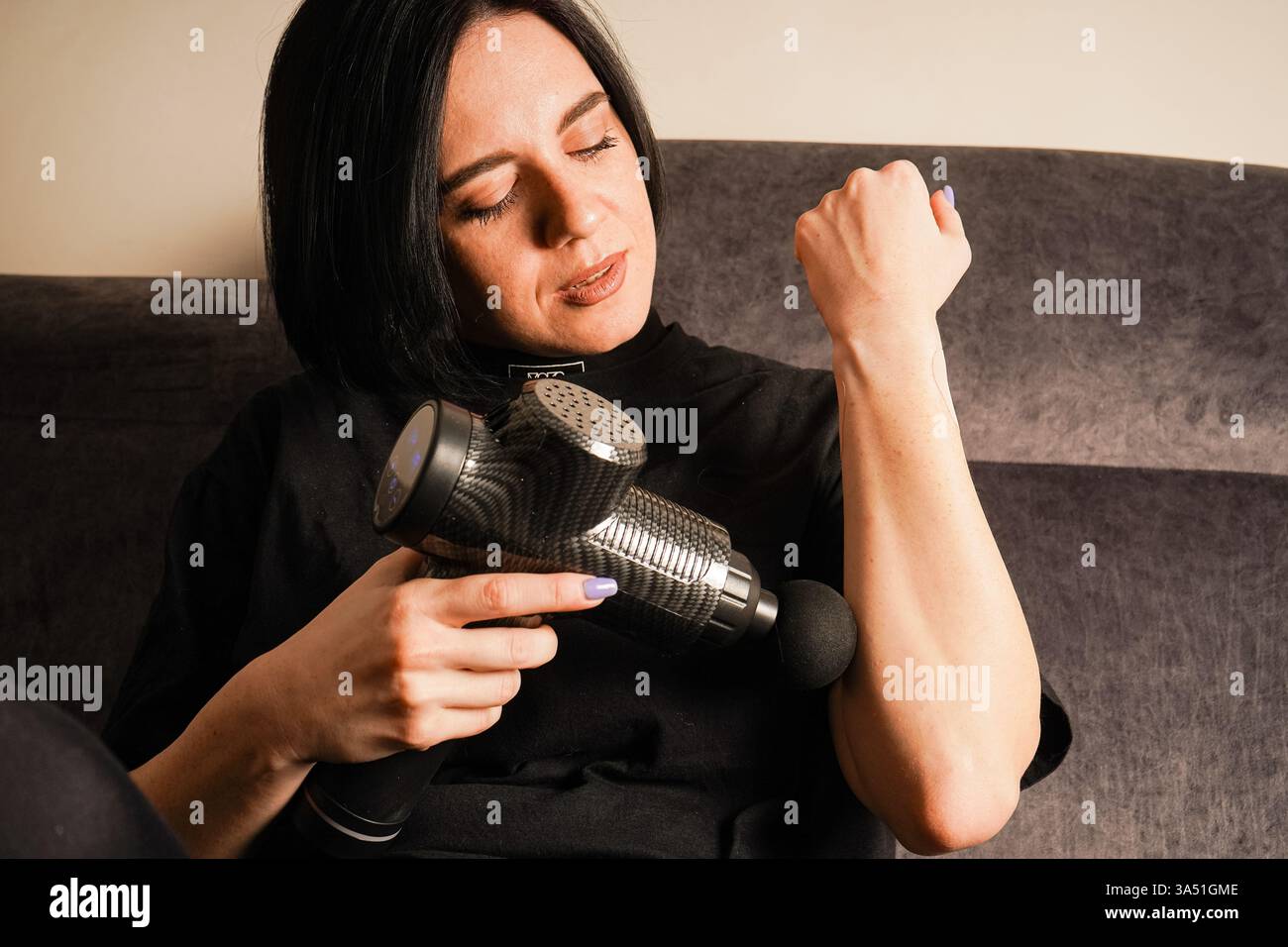 Sitting on a sofa, a woman massages her thigh using a handheld massage ...