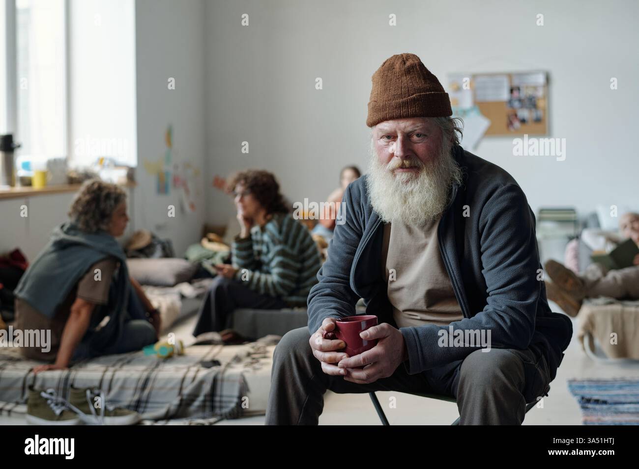 Senior man with grey beard holding mug and looking at camera while sitting in crisis camp for ...