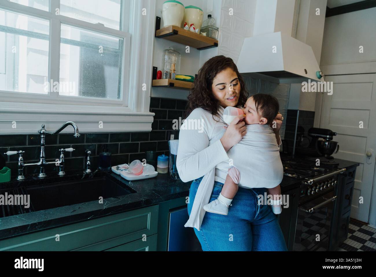 Hispanic mother wearing carrier feeding her baby with milk on bottle ...