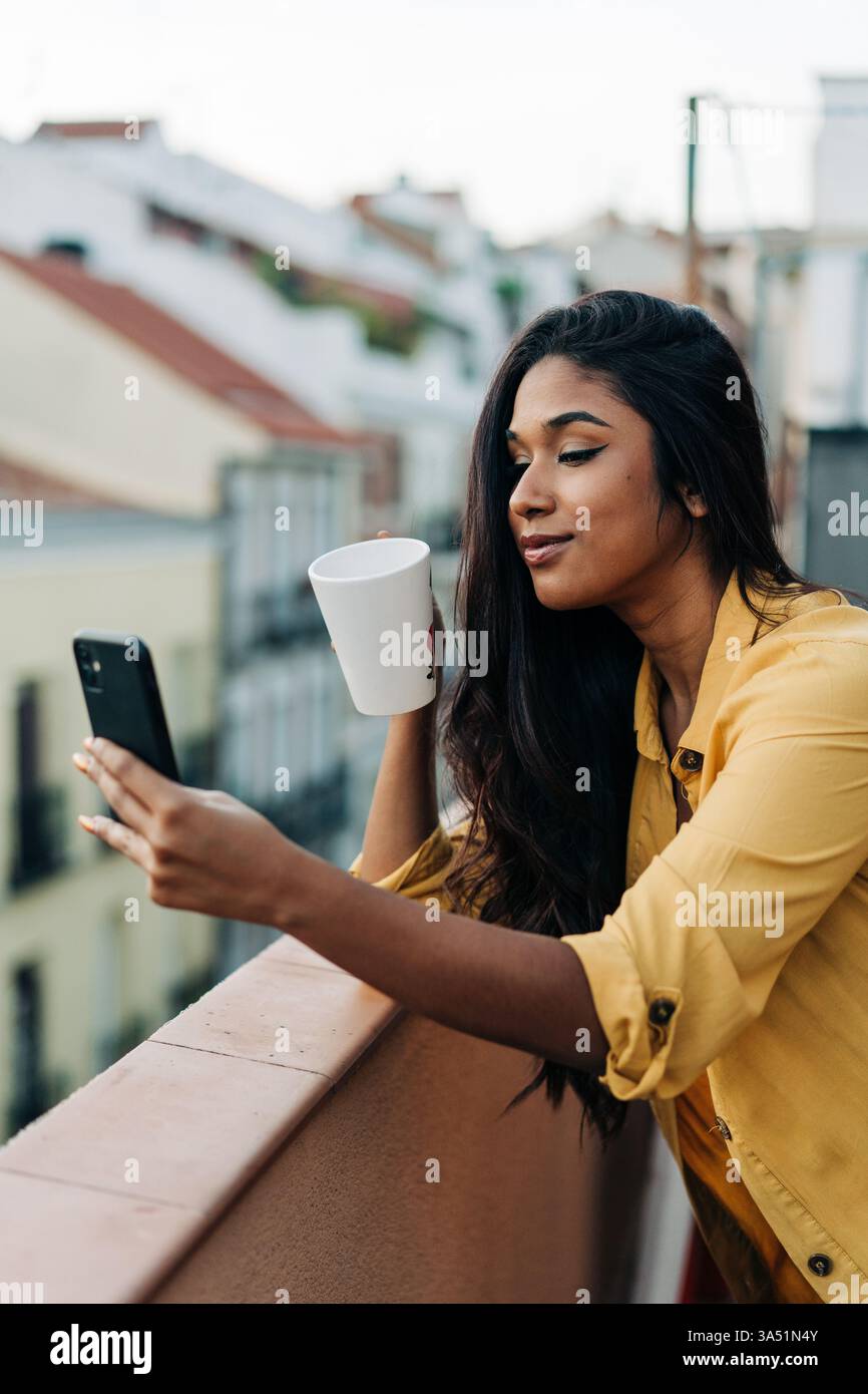 Hispanic female enjoying coffee and using smartphone while leaning on ...