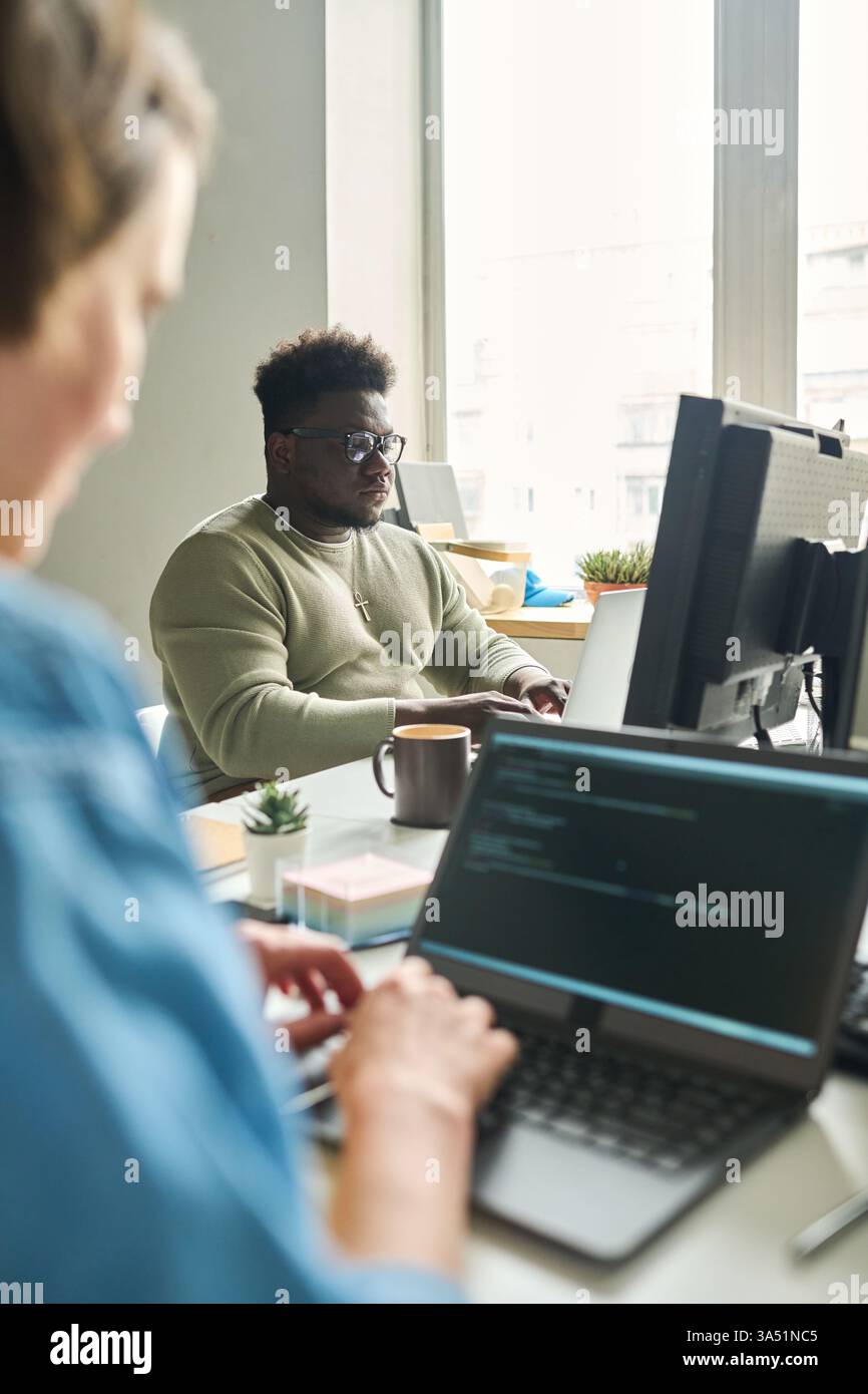 Serious Black male programmer wearing eyeglasses using laptop sitting at table with caucasian ...