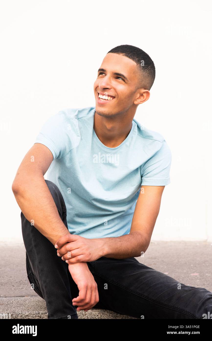 Arab male leaning arm on knee sitting on sidewalk against white wall ...