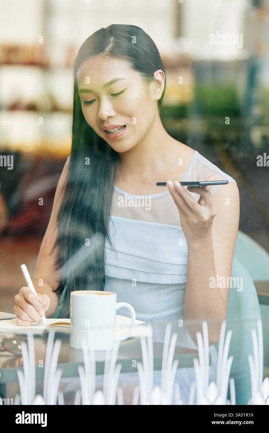 Young Asian woman working at cafe table, taking notes in planner and ...