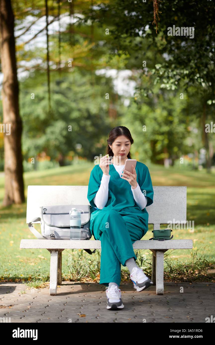 Nurse in scrubs sits on a park bench during the day, eating a snack and ...