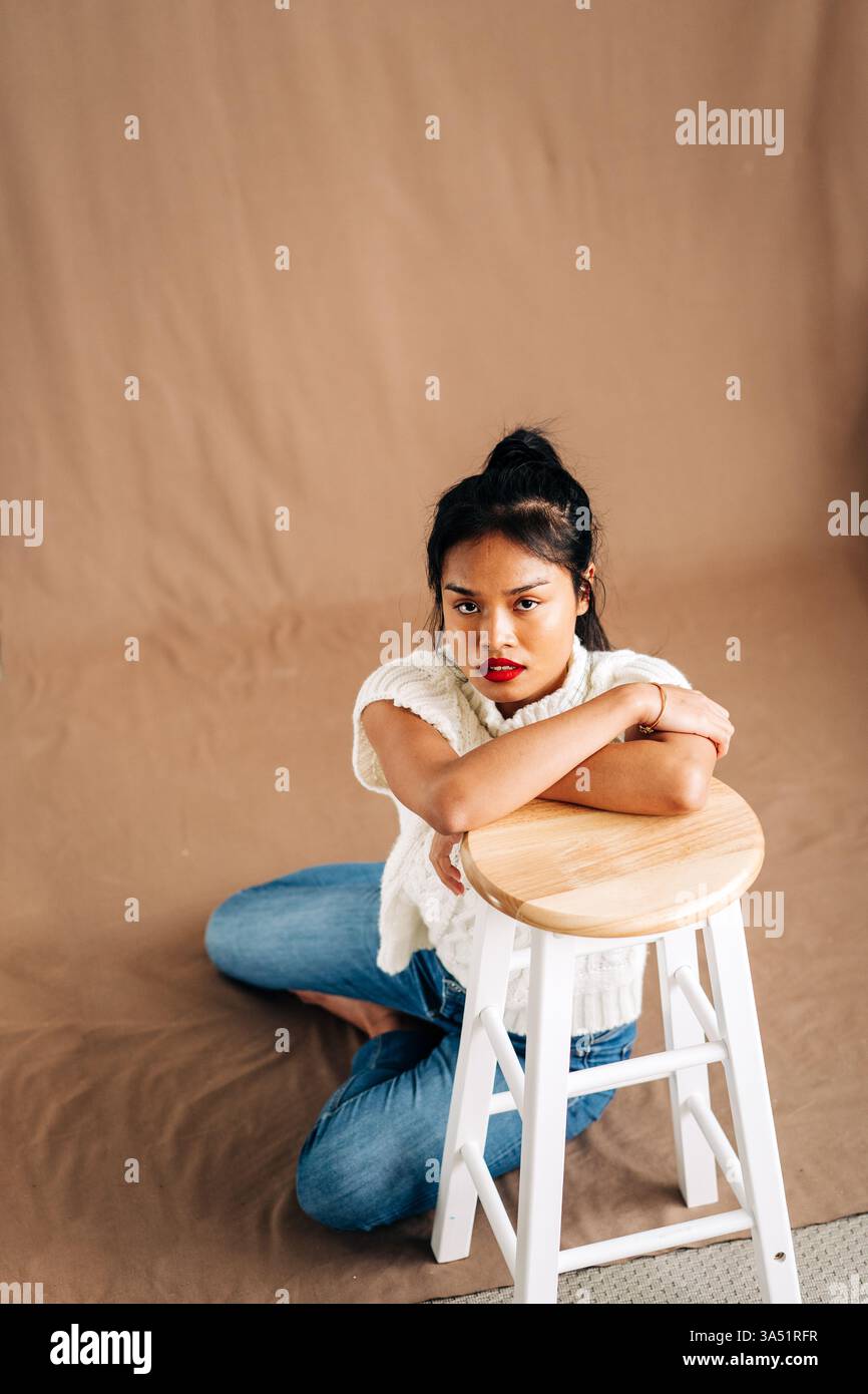 Asian woman sitting on the floor leaning on wooden stool in studio ...