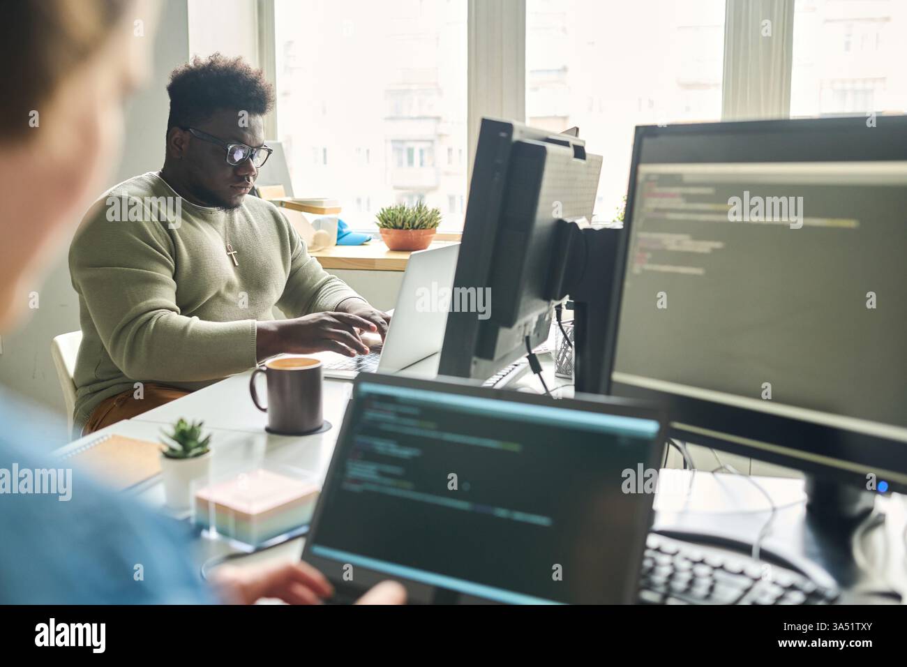 Serious Black male programmer wearing eyeglasses using laptop sitting at table with caucasian ...