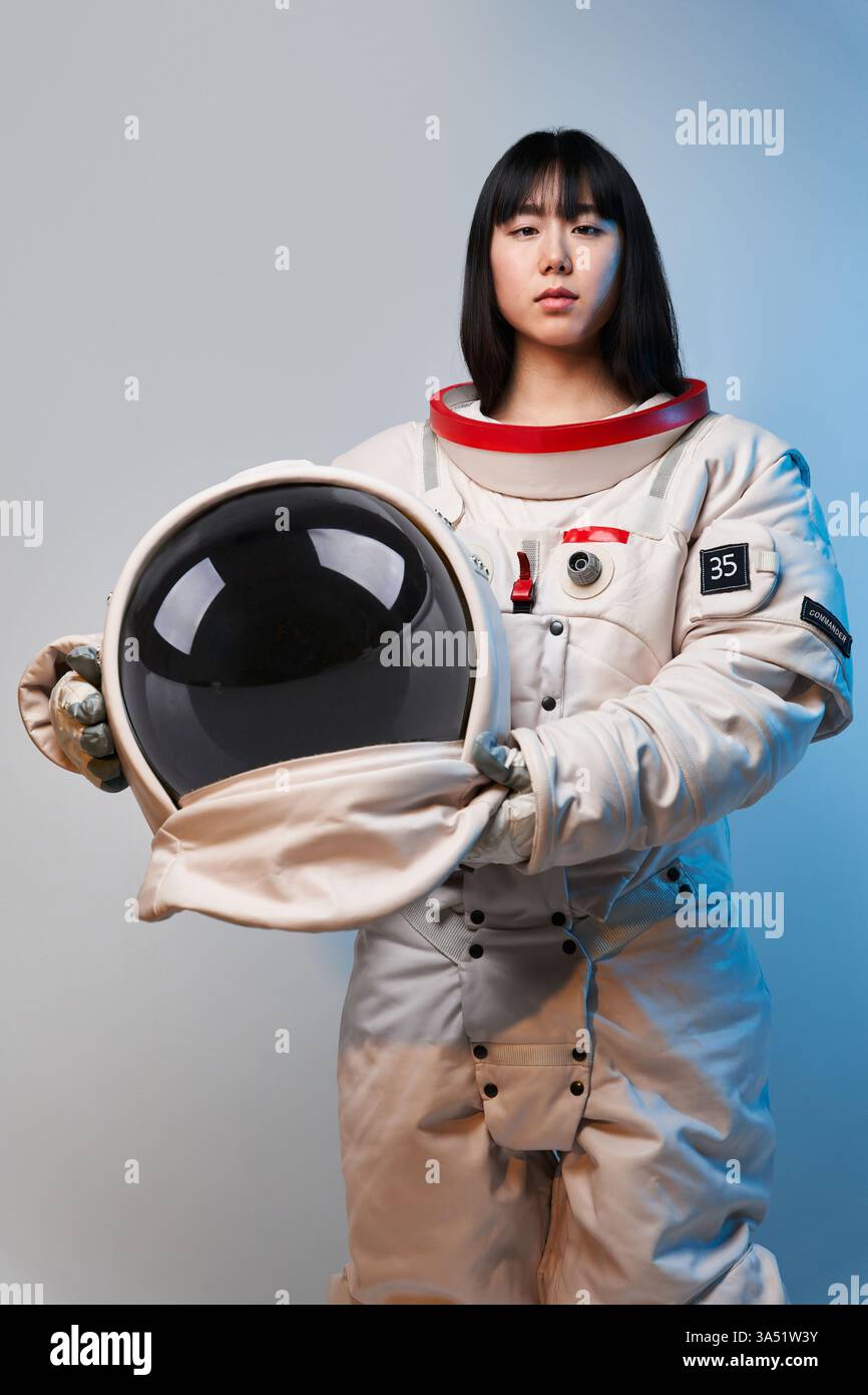 Serious Asian woman wearing spacesuit holding helmet standing with blue ...