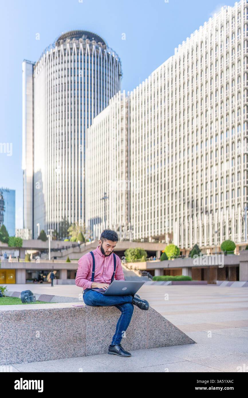 Bearded Asian man using laptop sitting with crossed legs on concrete ...