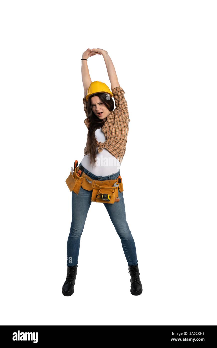 A female construction worker in a yellow hard hat and tool belt stretches her arms overhead ...