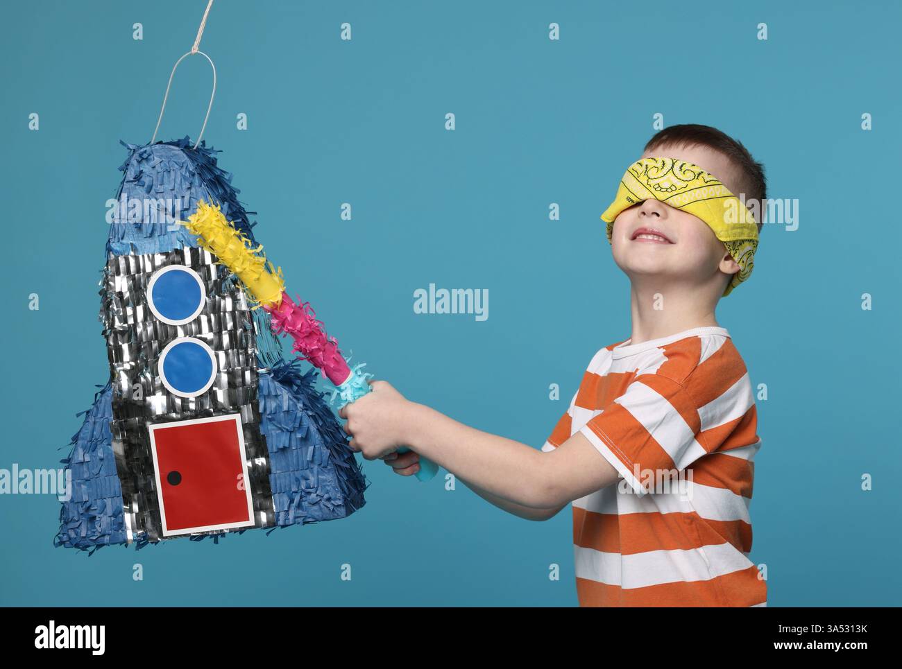 Happy boy with tied eyes breaking rocket shaped pinata on light blue ...