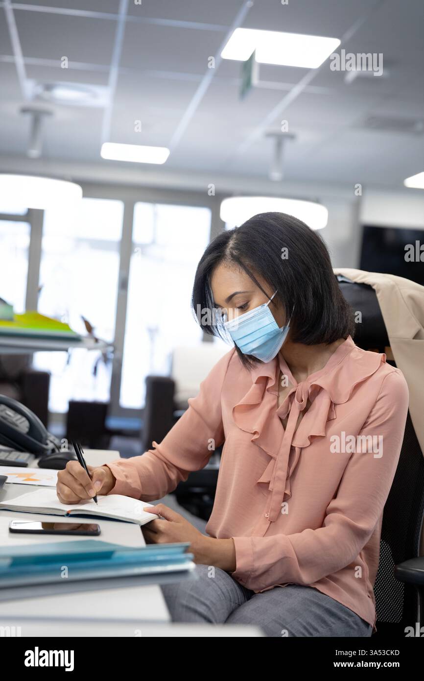 Professional woman wearing mask writing notes at office desk, focused ...