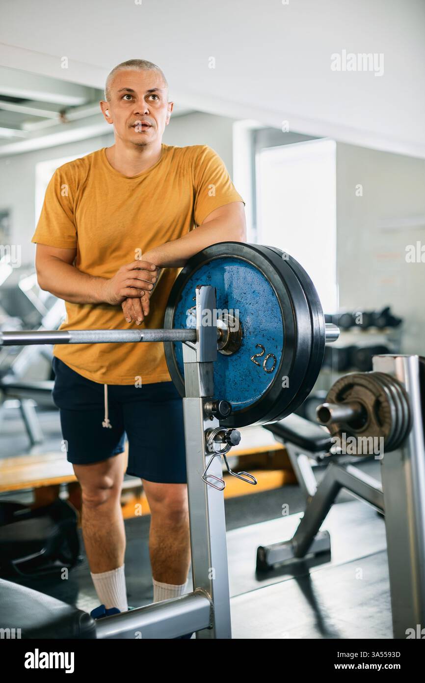 A senior man taking a break after lifting weights in a gym, leaning on ...