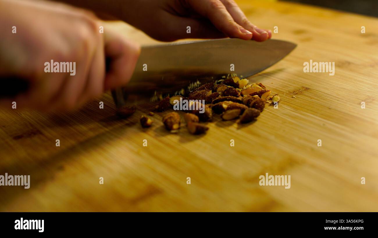 Hands chopping up nuts with a sharp kitchen knife on a wooden counter ...
