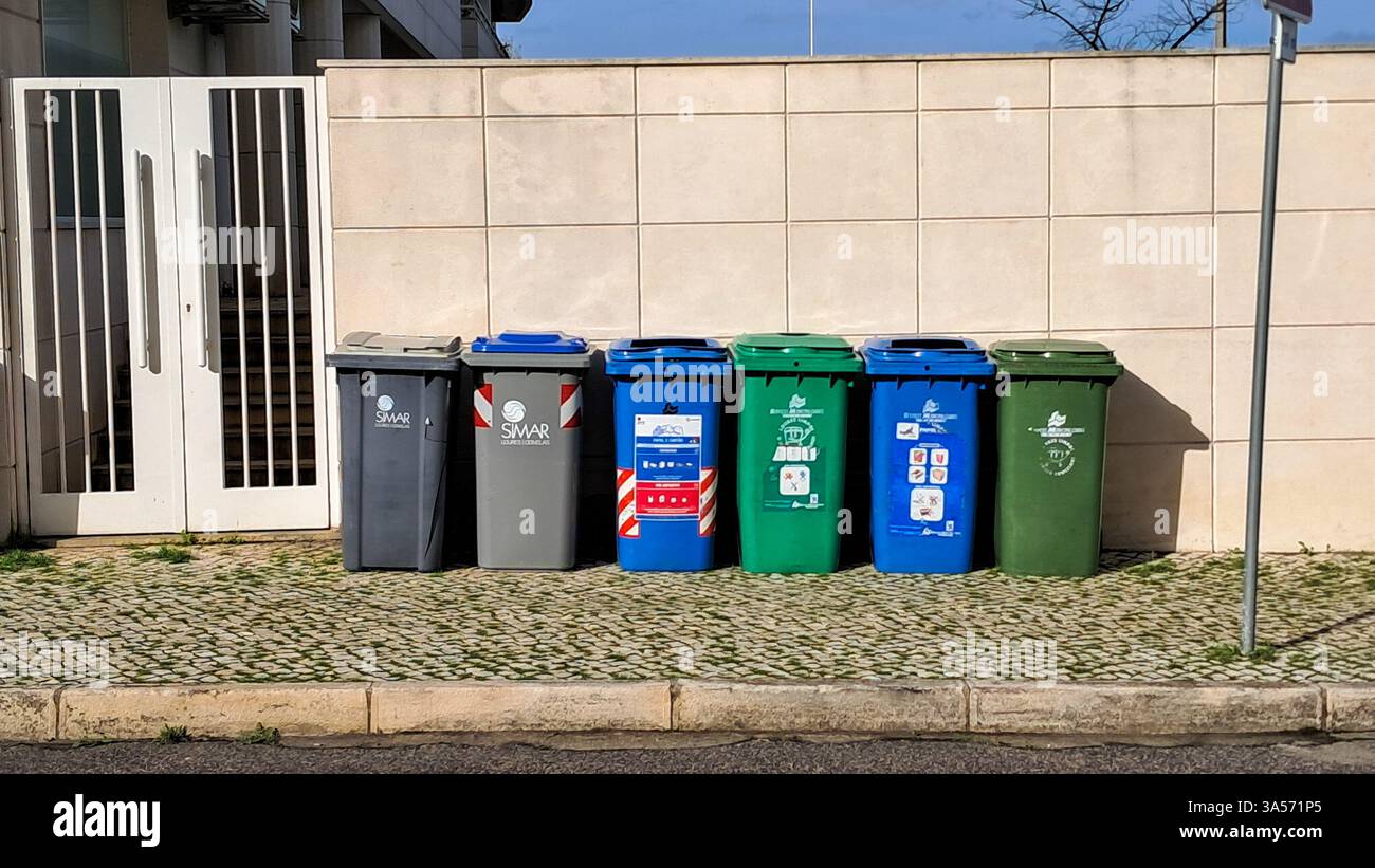Different colored waste bins stand on the sidewalk next to a tiled wall ...
