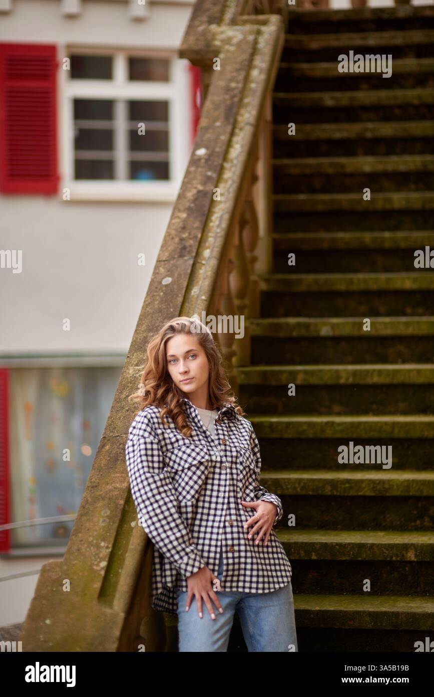 Stylish young woman posing on an old mossy staircase in a European town with red window shutters ...