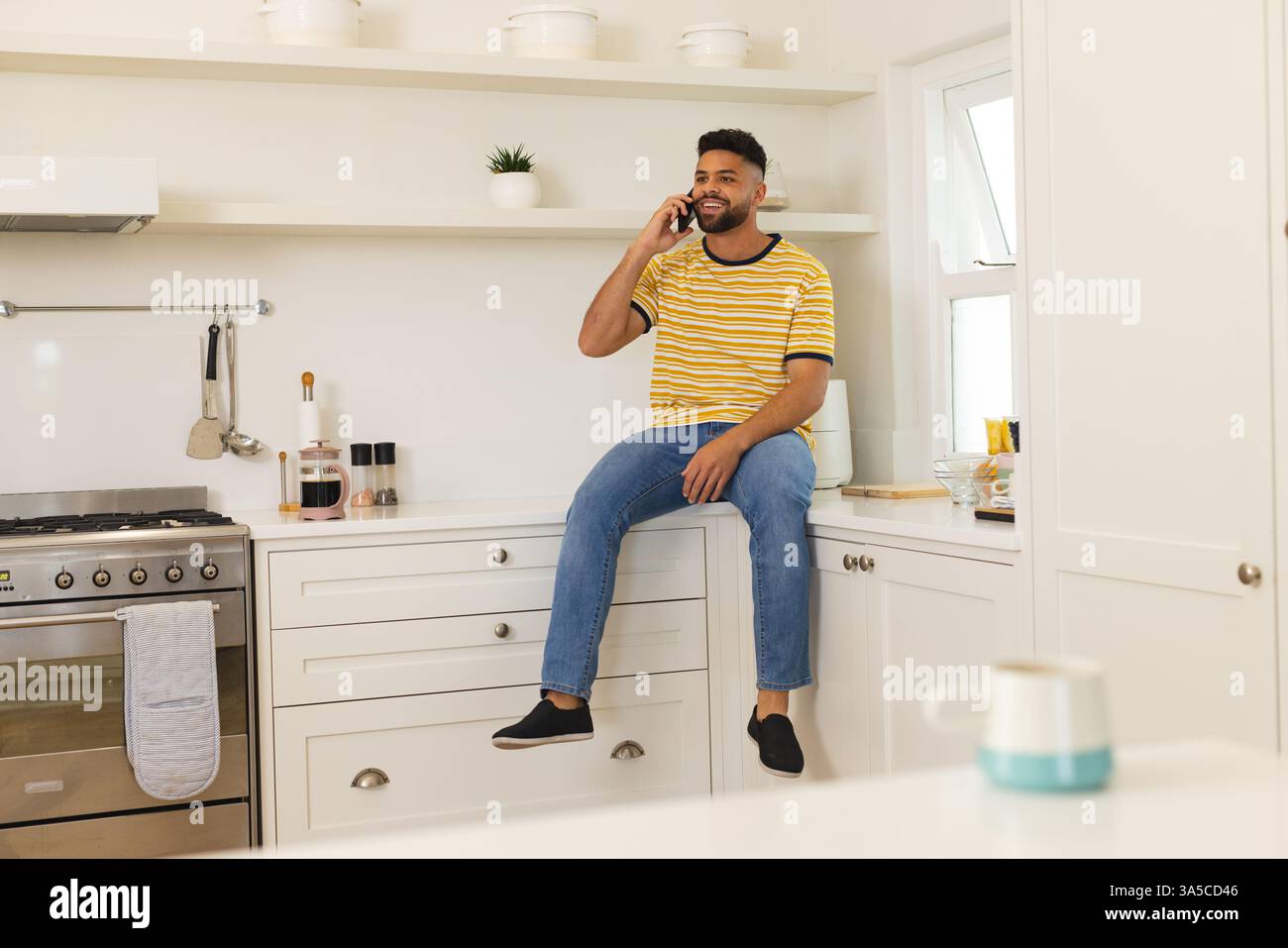 Young man sitting on kitchen counter, talking on smartphone, enjoying ...