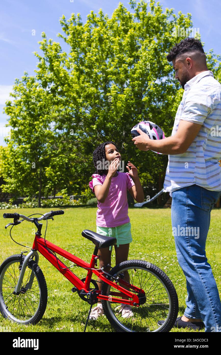 Father handing bicycle helmet to daughter in park, preparing for bike ...