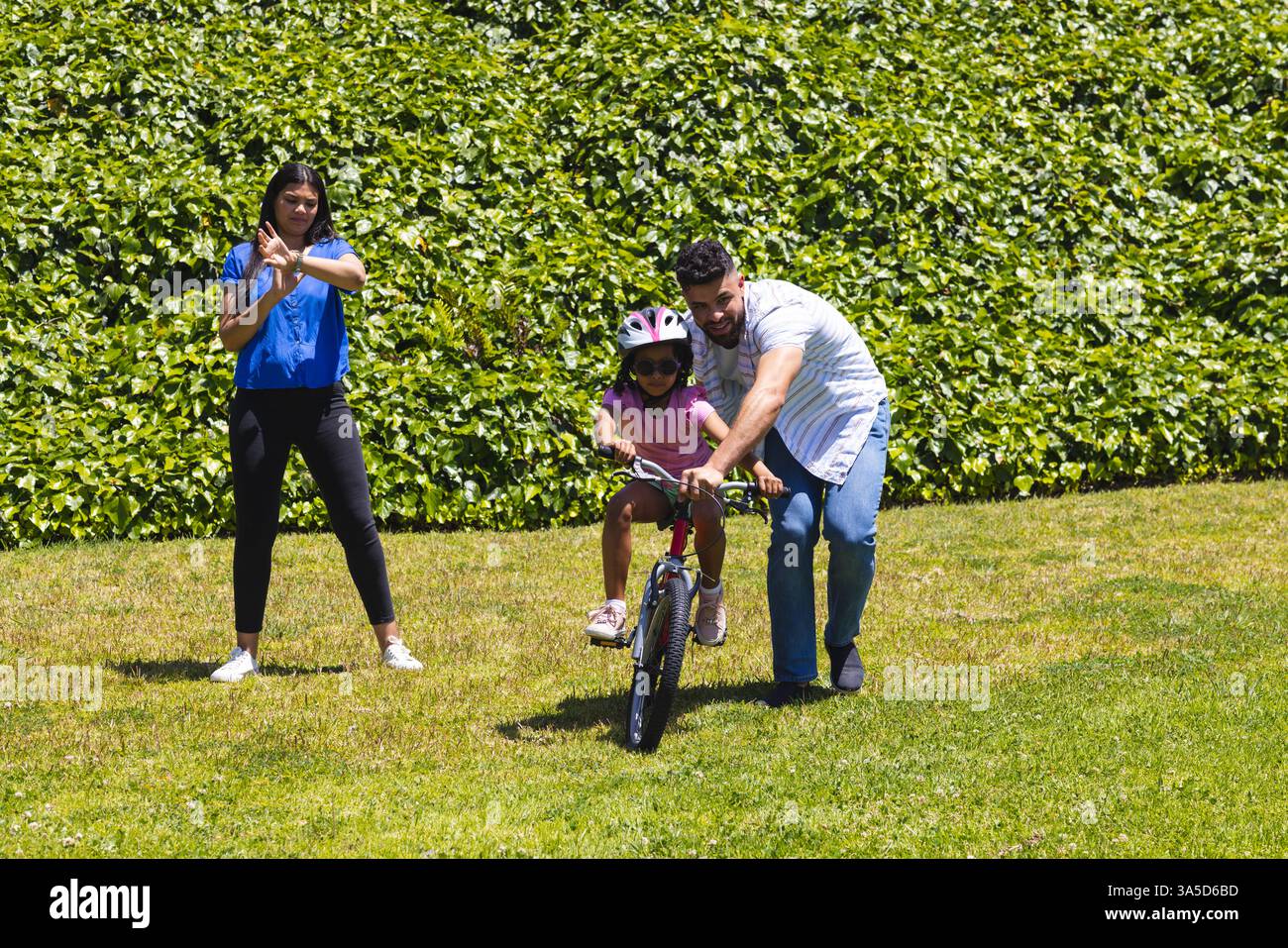Father teaching daughter to ride bicycle while mother cheering in park ...