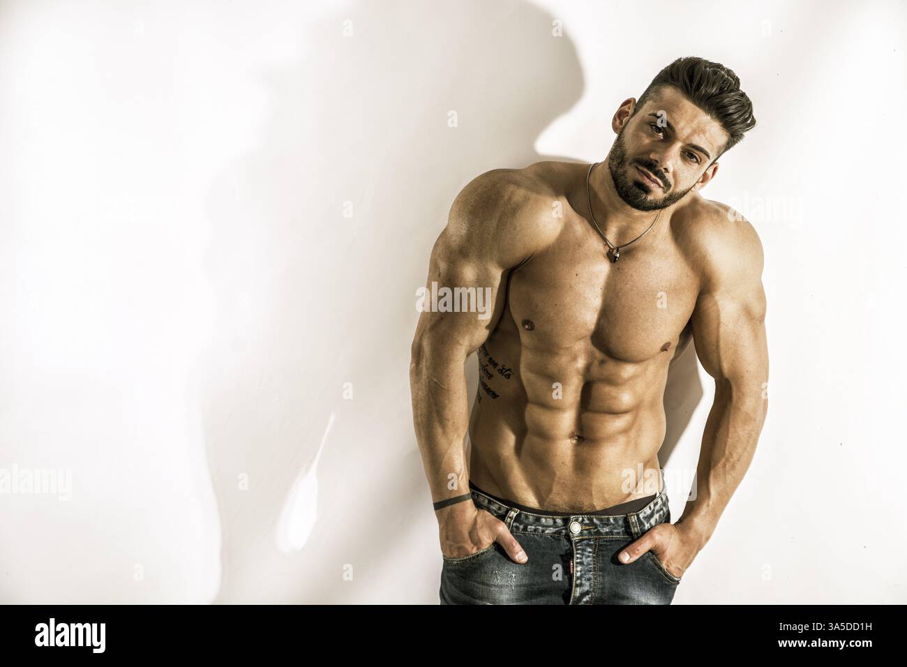 Muscular bodybuilder leaning against white wall, looking at camera ...
