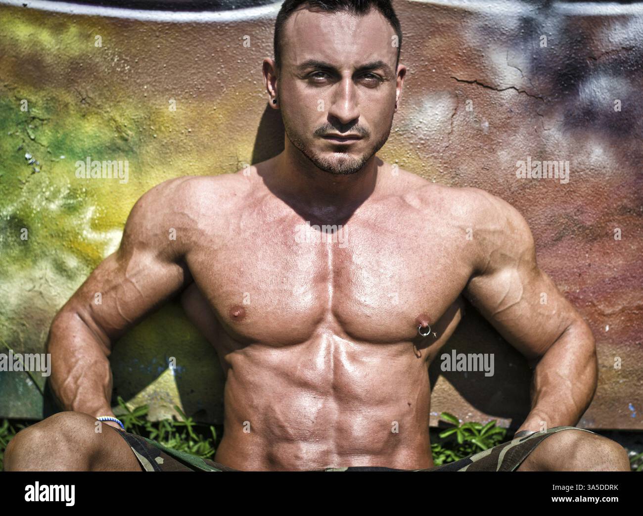 Muscular bodybuilder sitting against colorful wall showing ripped pecs ...