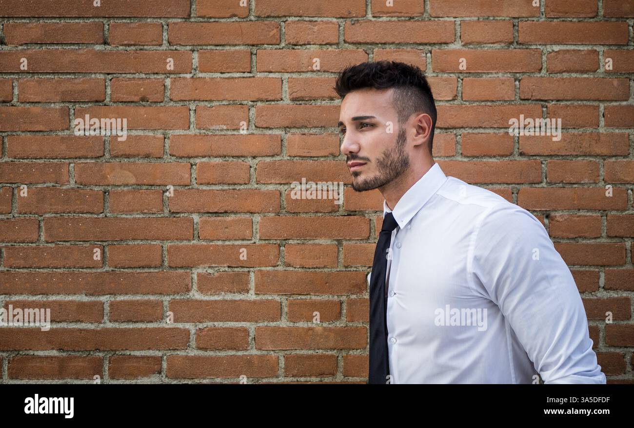 Attractive young man standing against brick wall, looking away. Half ...