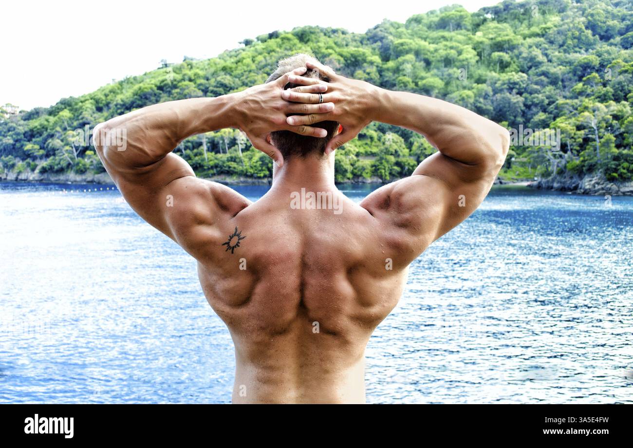 Muscular bodybuilder facing the sea with hands behind his head showing ...