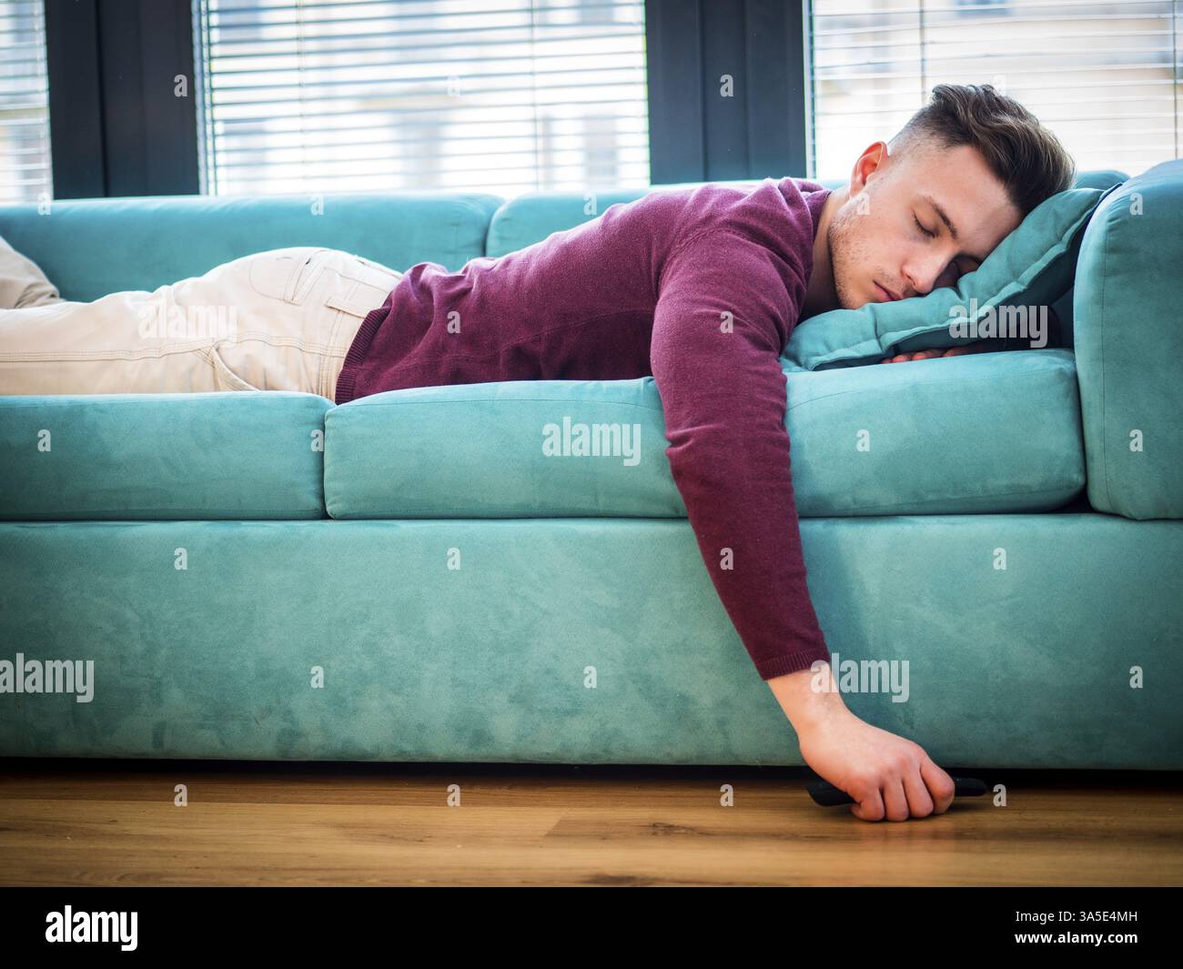 Young bored man lying on sofa watching television changing the channel ...
