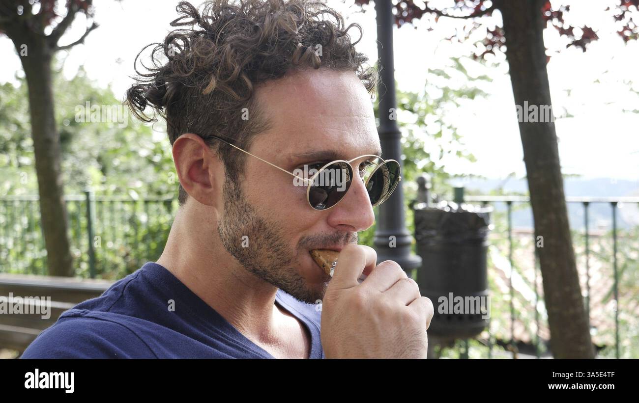 Handsome young man eating cereal bar, looking away sitting on terrace ...