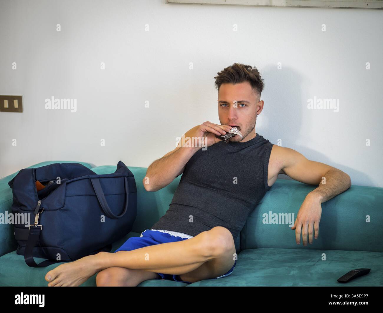 Young muscular man eating protein bar indoor at home sitting on a couch ...