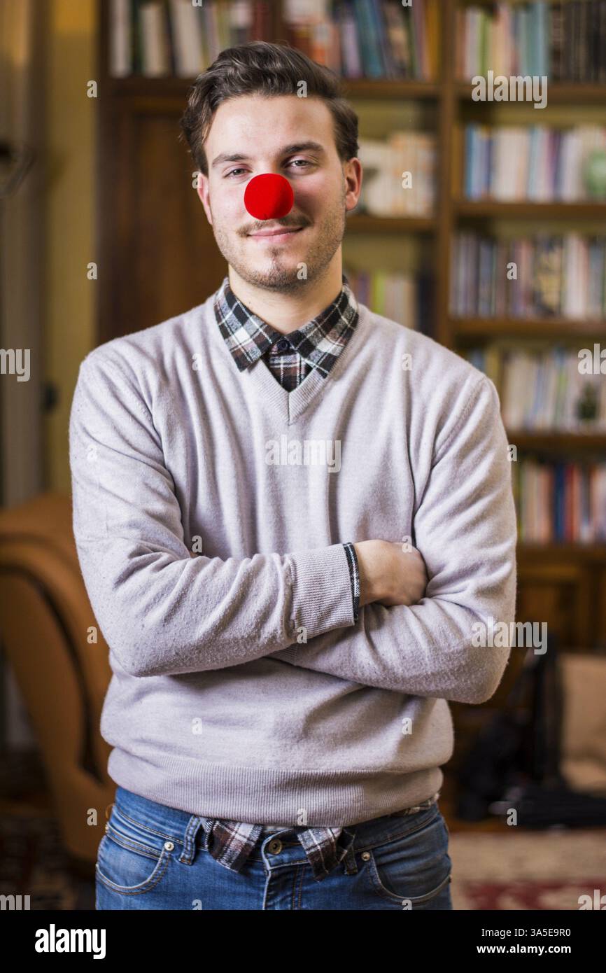 Handsome young man smiling with red clown nose, standing in a living ...