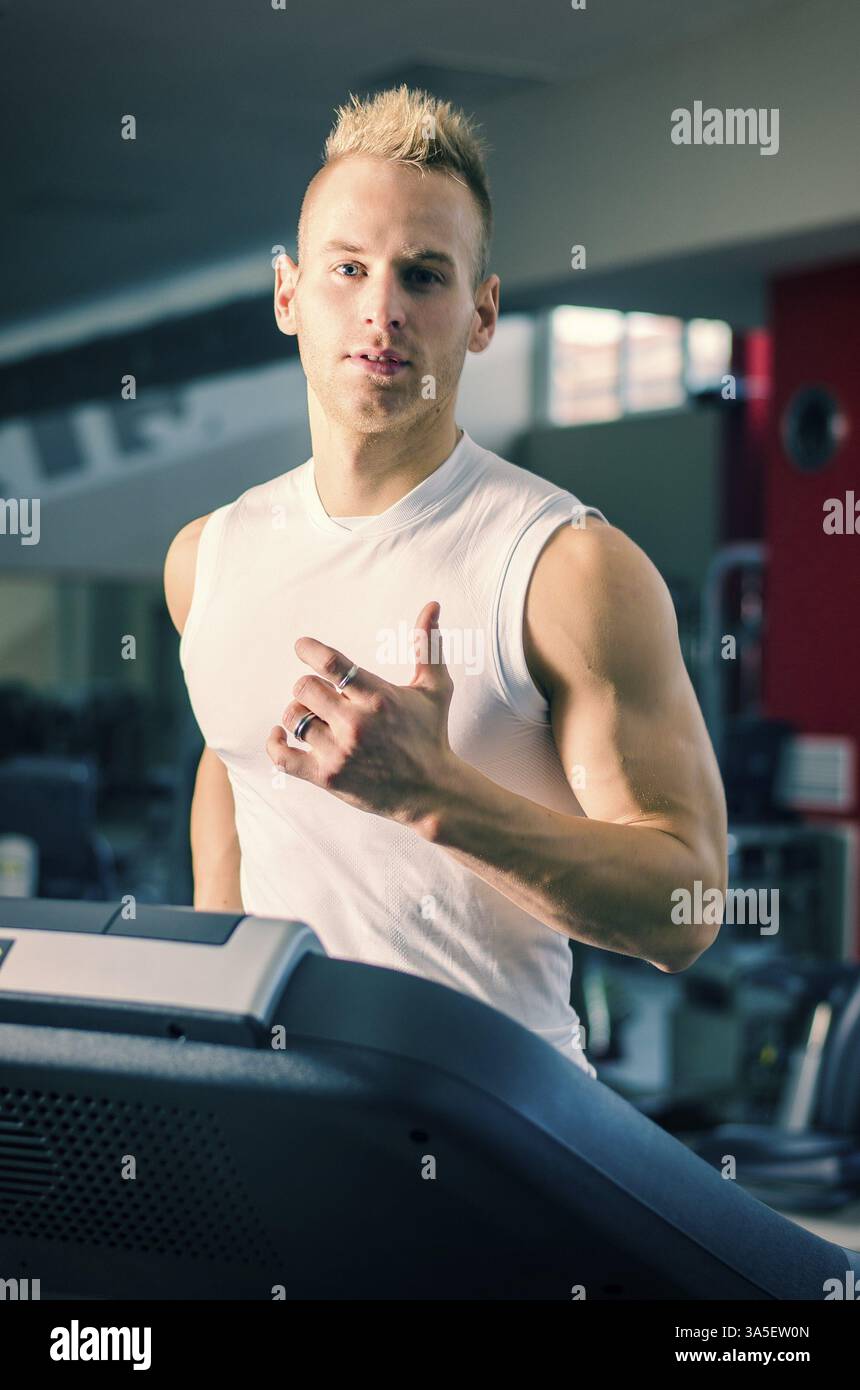 Blond attractive young man running on treadmill, looking at camera ...
