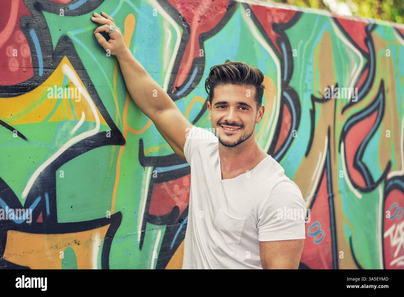 Attractive muscle man leaning on colorful graffiti wall, wearing white ...