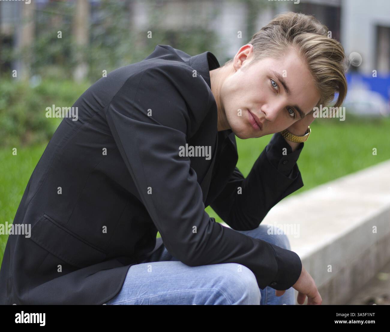 Serious or sad blond young man in jeans and jacket, sitting outdoors ...