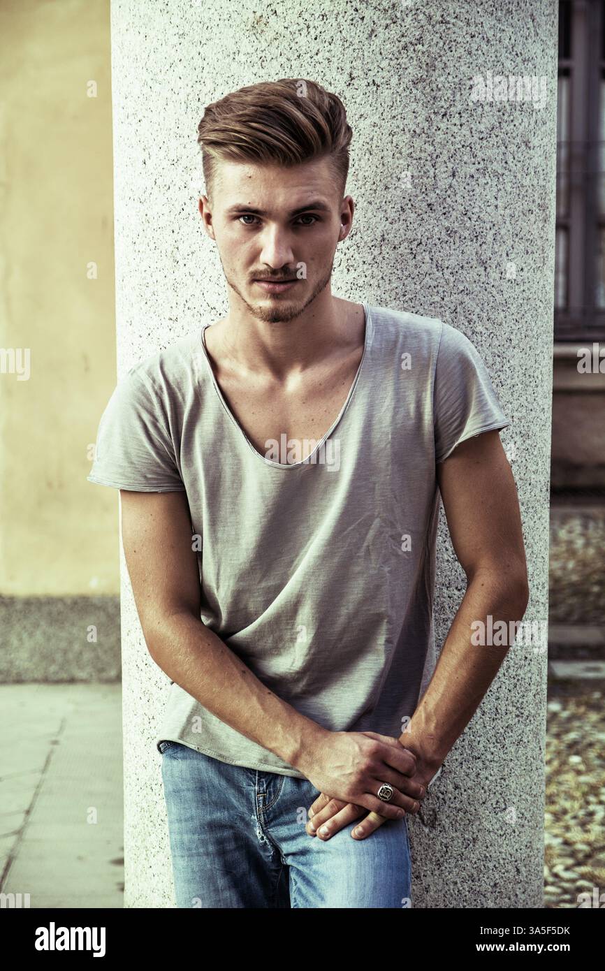 Handsome blond young man, blue eyes, leaning against white, rough stone ...