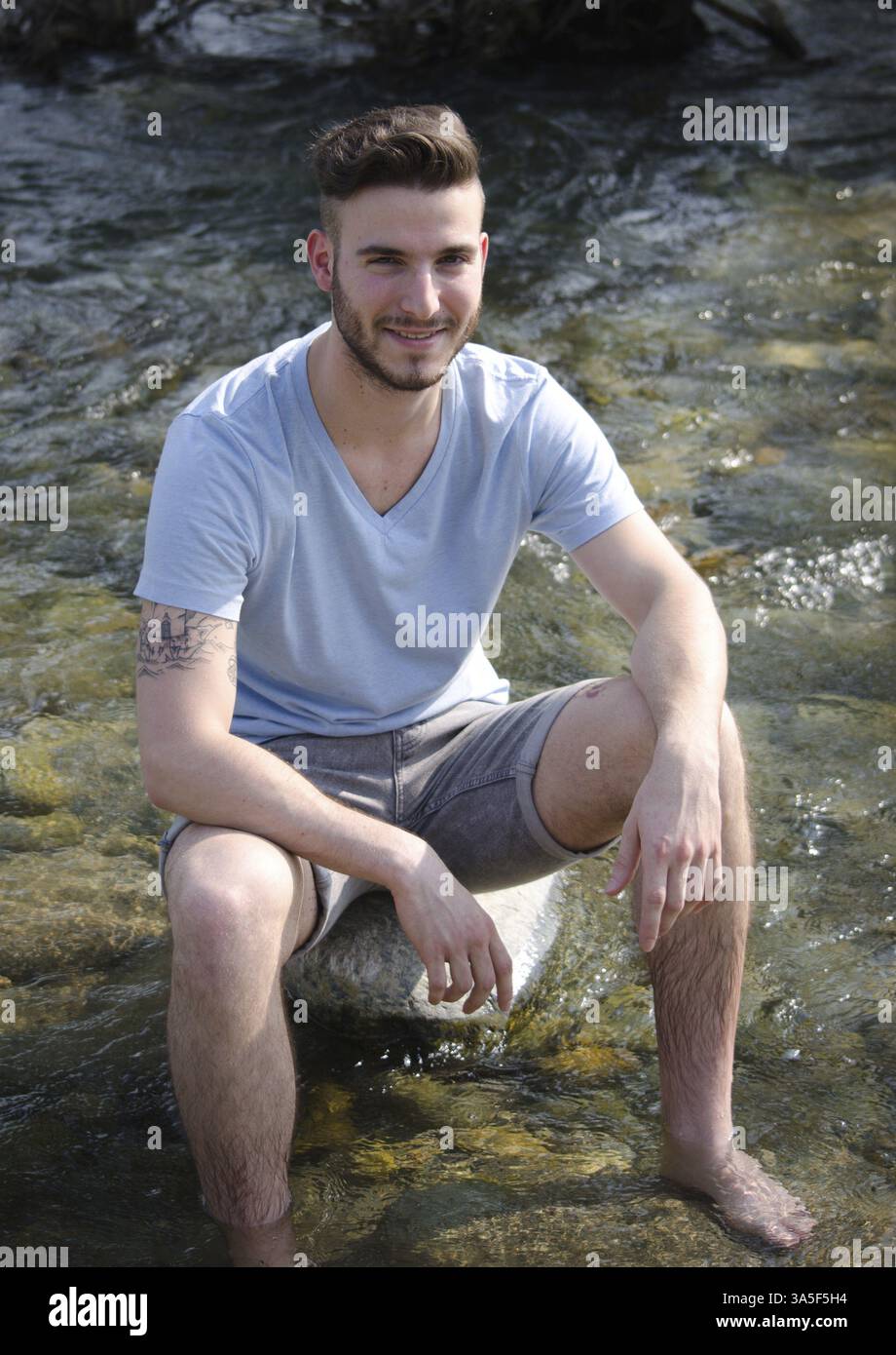 Attractive young man sitting on rock in a river, smiling with feet in ...