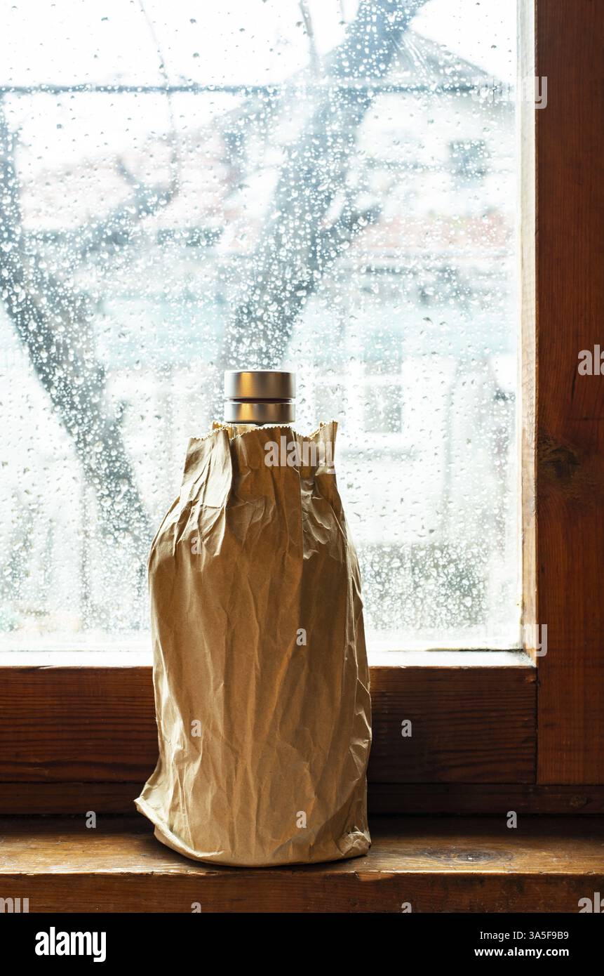 Bottle with alcohol in a brown pack paper placed to the window at rainy ...